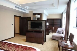Quarto de hotel com cama, televisão em aparador de madeira, uma cadeira, mesa pequena e estante.