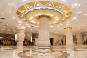 Um hall amplo com grandes colunas, um lustre circular central e piso de brilho intenso.