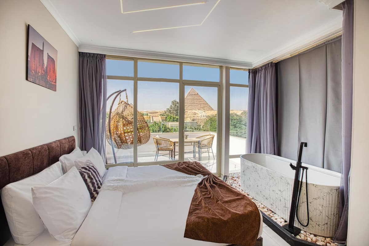 Quarto de hotel com uma cama, vista da pirâmide e varanda com mesa e cadeiras.