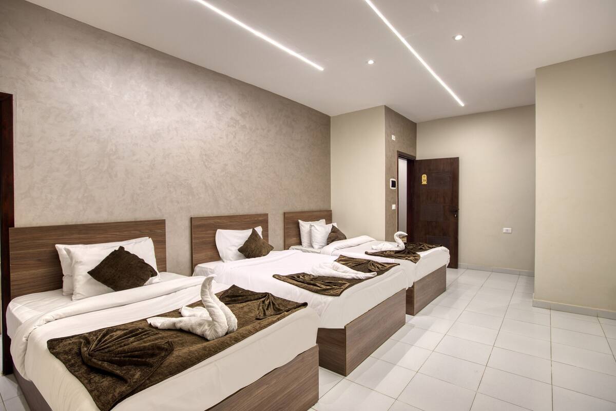 Quarto de hotel com três camas, cada uma com cabeceira de madeira, roupa de cama branca e mantas marrons. O quarto possui piso de azulejos e uma parede lisa com uma porta.