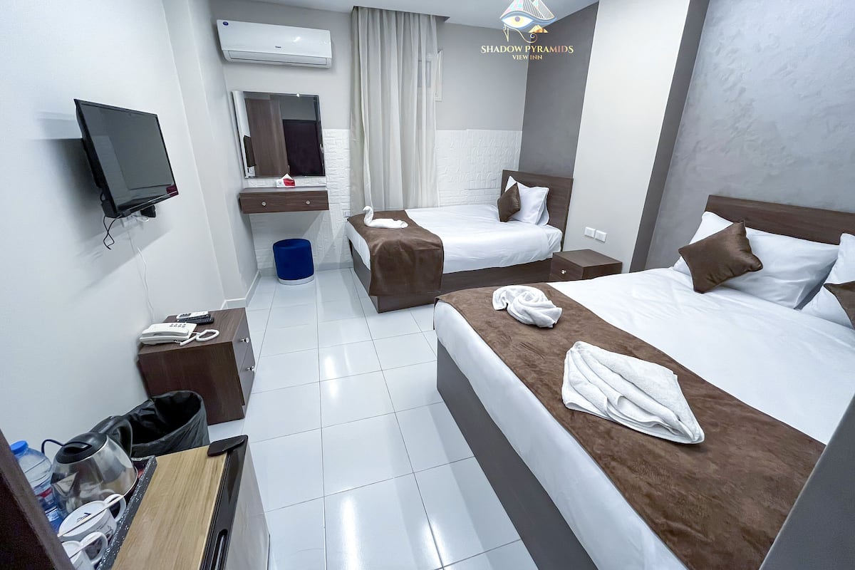 Quarto de hotel com duas camas, uma televisão, uma secretária com computador, um cesto de lixo e um dispensador de água.