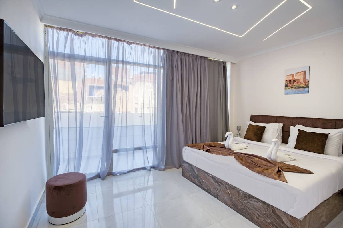 Quarto de hotel com uma cama grande, um banquinho redondo, cortinas translúcidas e um quadro na parede.