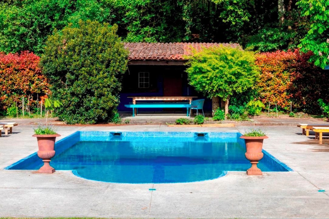 Uma piscina com borda azulejada, rodeada por vegetação e um pequeno edifício com telhado em azulejos.