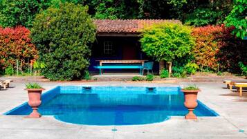 Uma piscina com borda azulejada, rodeada por vegetação e um pequeno edifício com telhado em azulejos.
