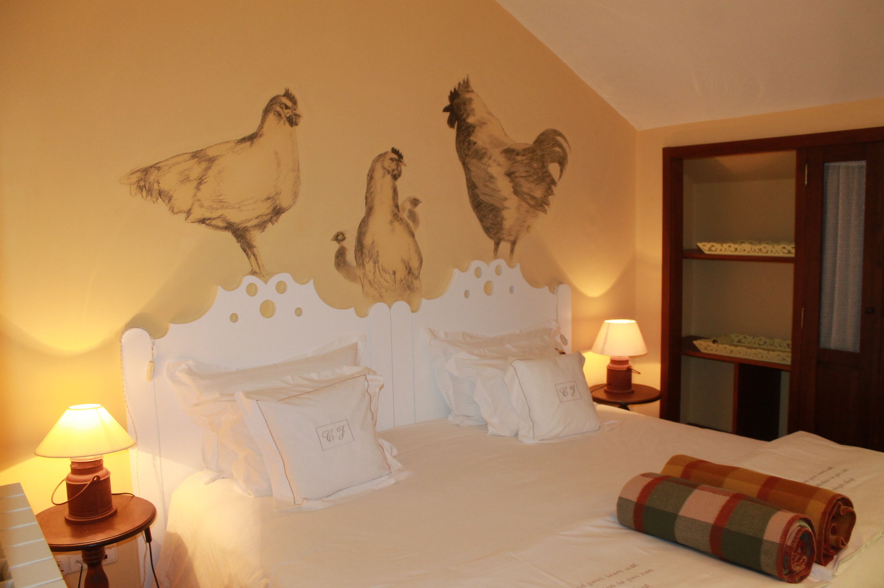 Quarto com cama, duas luminárias de cabeceira e um mural de galinhas na parede.