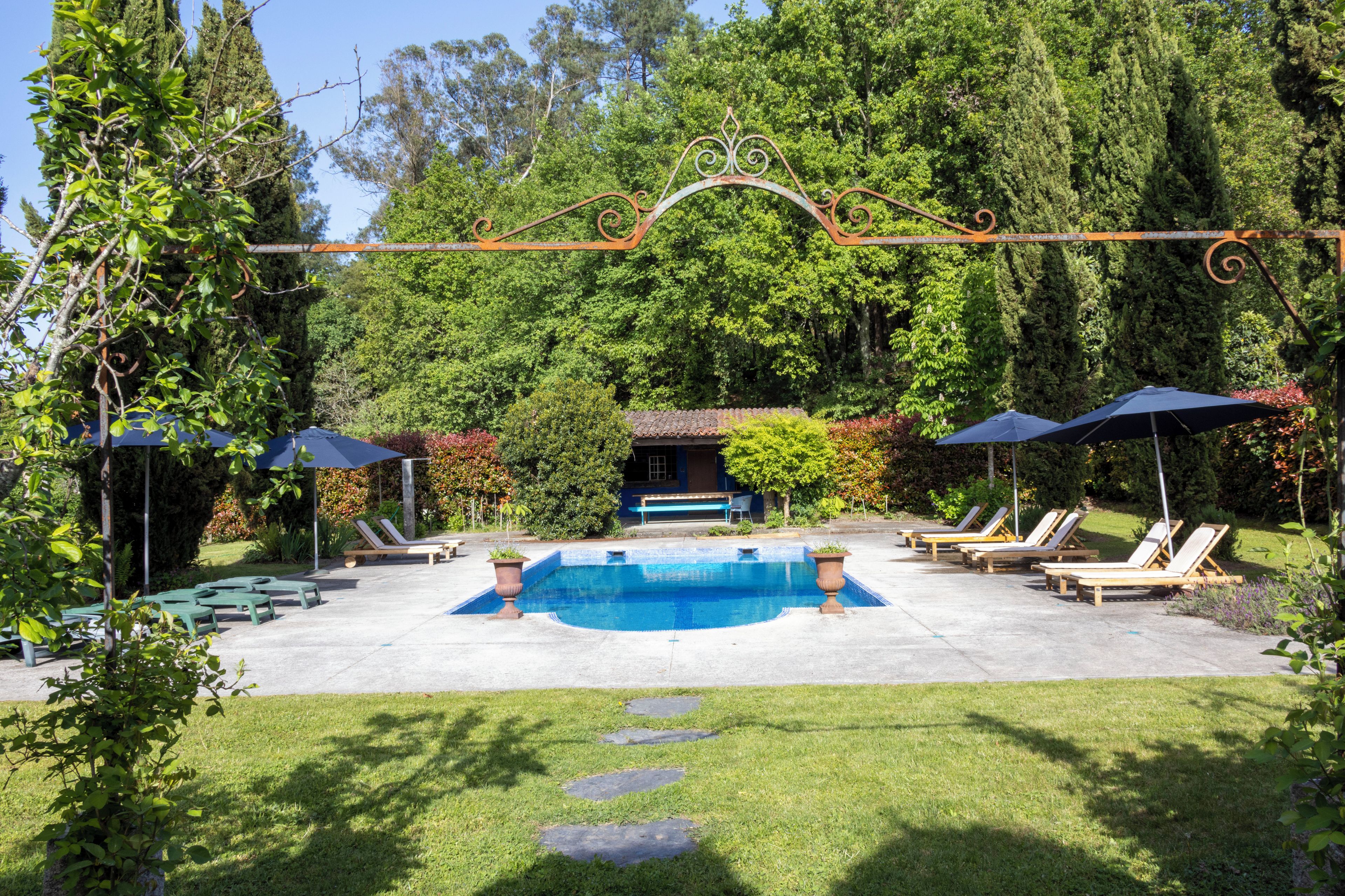 Piscina com espreguiçadeiras e guarda-sóis, cercada por árvores e um pequeno edifício.