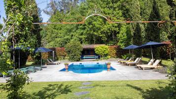 Piscina com espreguiçadeiras e guarda-sóis, cercada por árvores e um pequeno edifício.