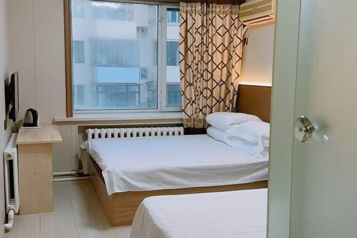 Quarto de hotel com duas camas, janela com vista para edifícios, radiador e escrivaninha com luminária.