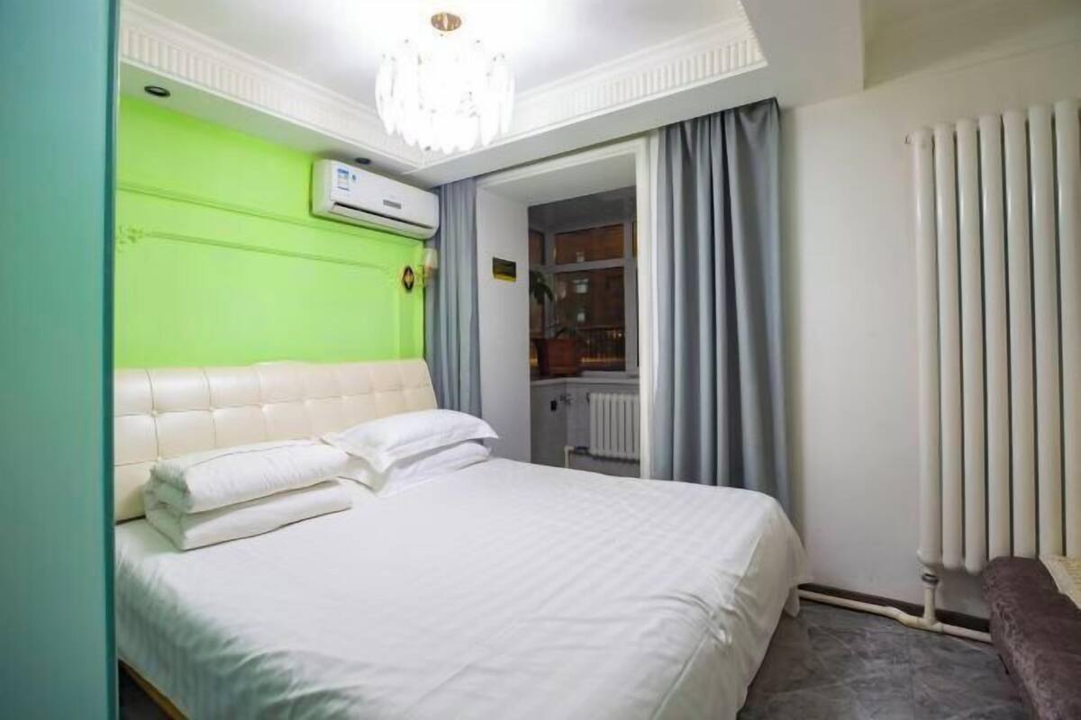 Quarto de hotel com cama, radiador, janela com cortinas e porta que dá para outro aposento.