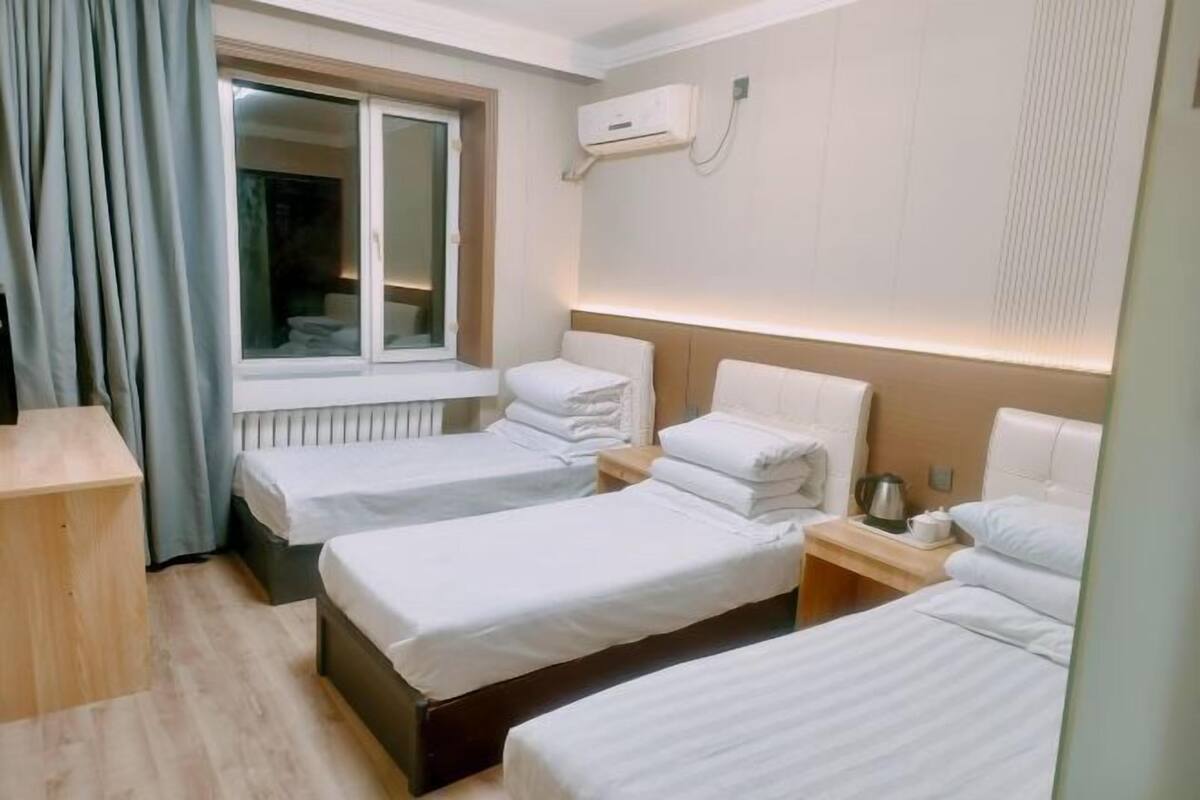 Quarto de hotel com duas camas, janela com cortinas, aparelho de ar condicionado, mesa de cabeceira com chaleira e pequena escrivaninha de madeira.