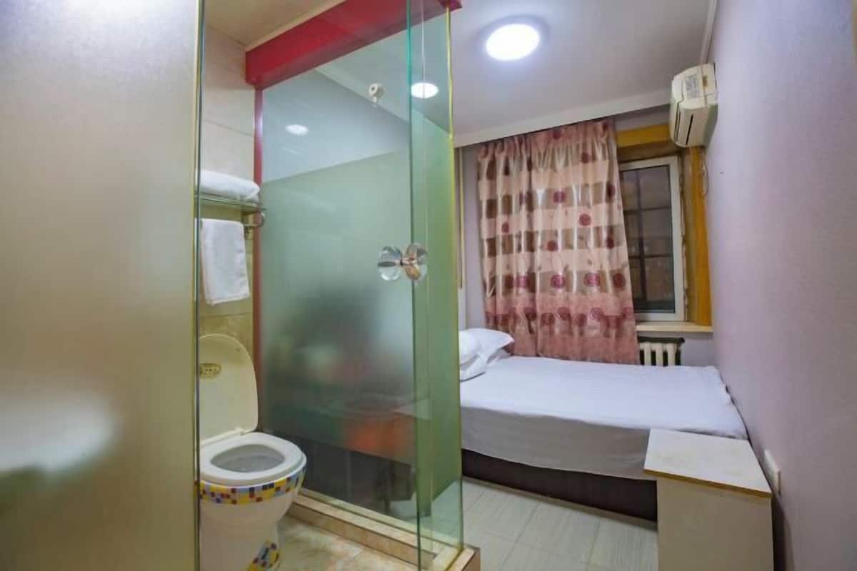 Quarto pequeno e compacto com chuveiro em box de vidro, uma cama de solteiro e vaso sanitário.