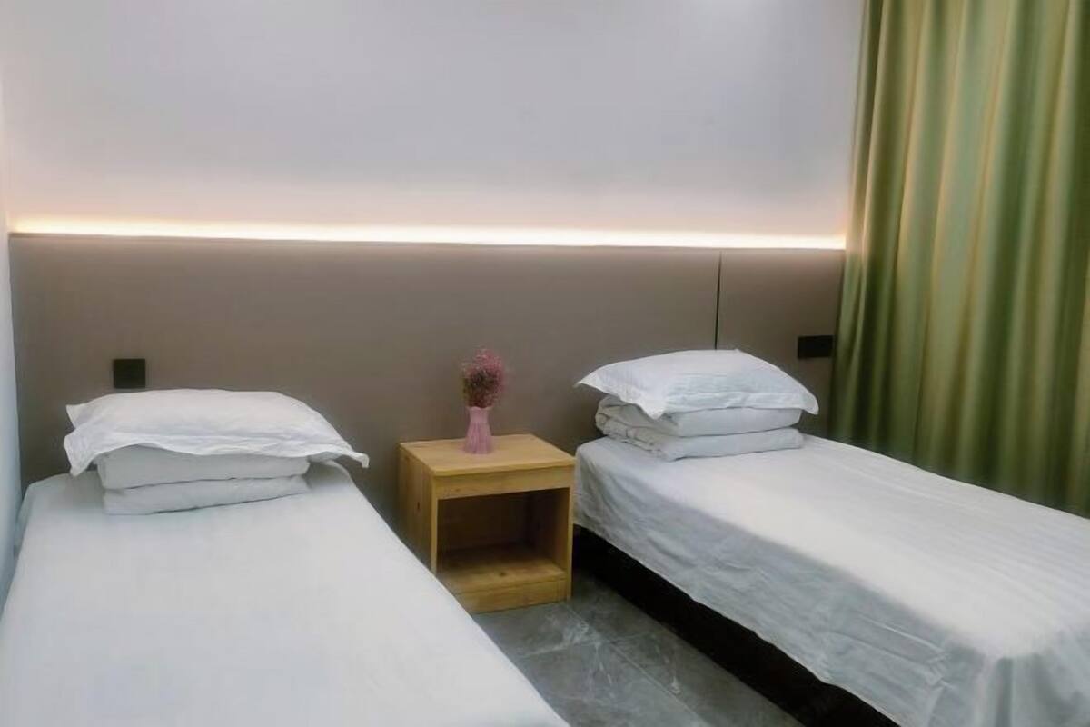 Quarto de hotel com duas camas de solteiro, uma mesinha de cabeceira de madeira com um vaso, e cortina verde.