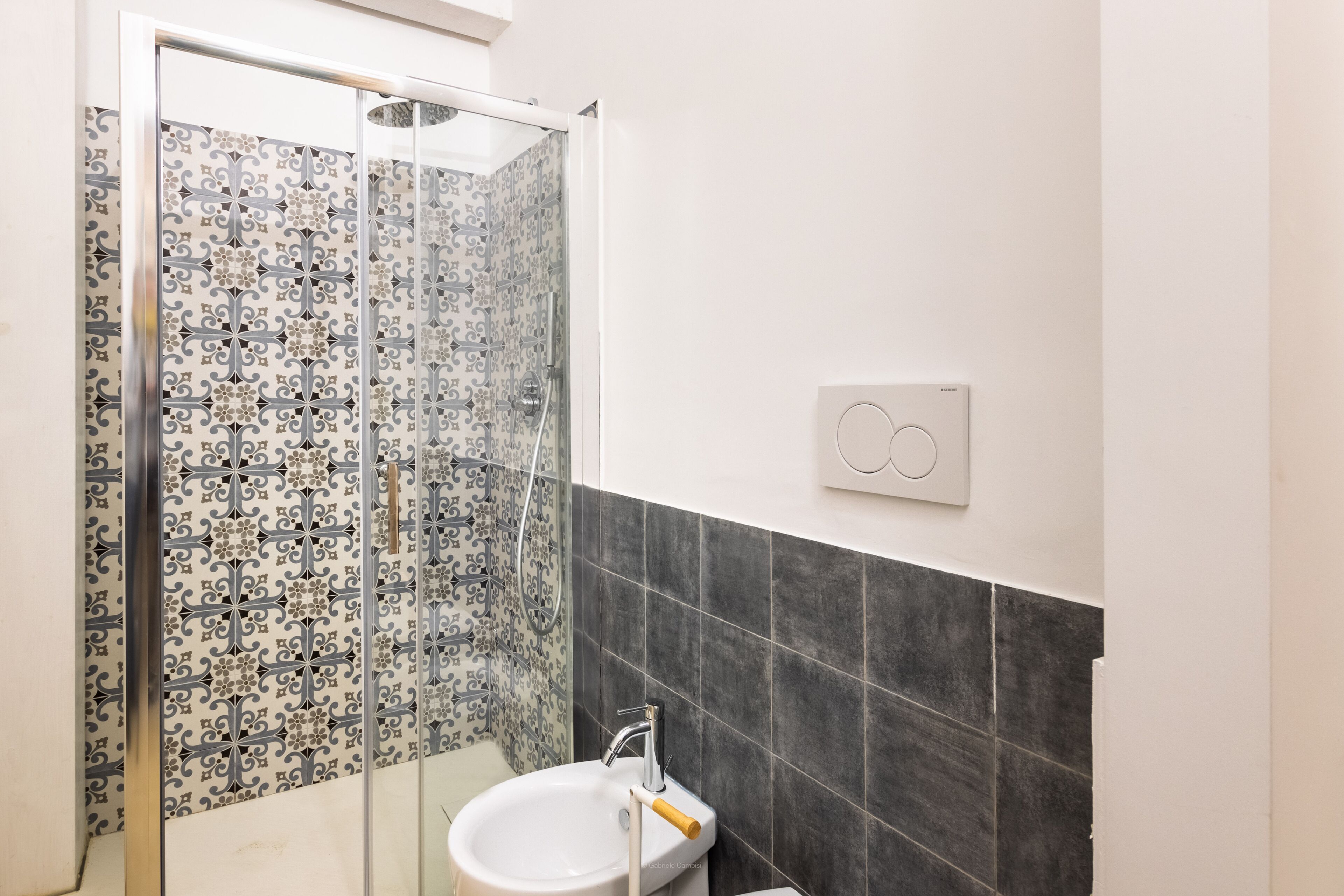 Casa de banho com cabine de duche em vidro, lavatório branco e azulejos estampados nas paredes.