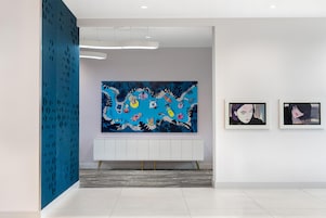 Uma galeria de arte moderna com uma grande pintura abstrata, um console branco e duas obras de arte emolduradas na parede.