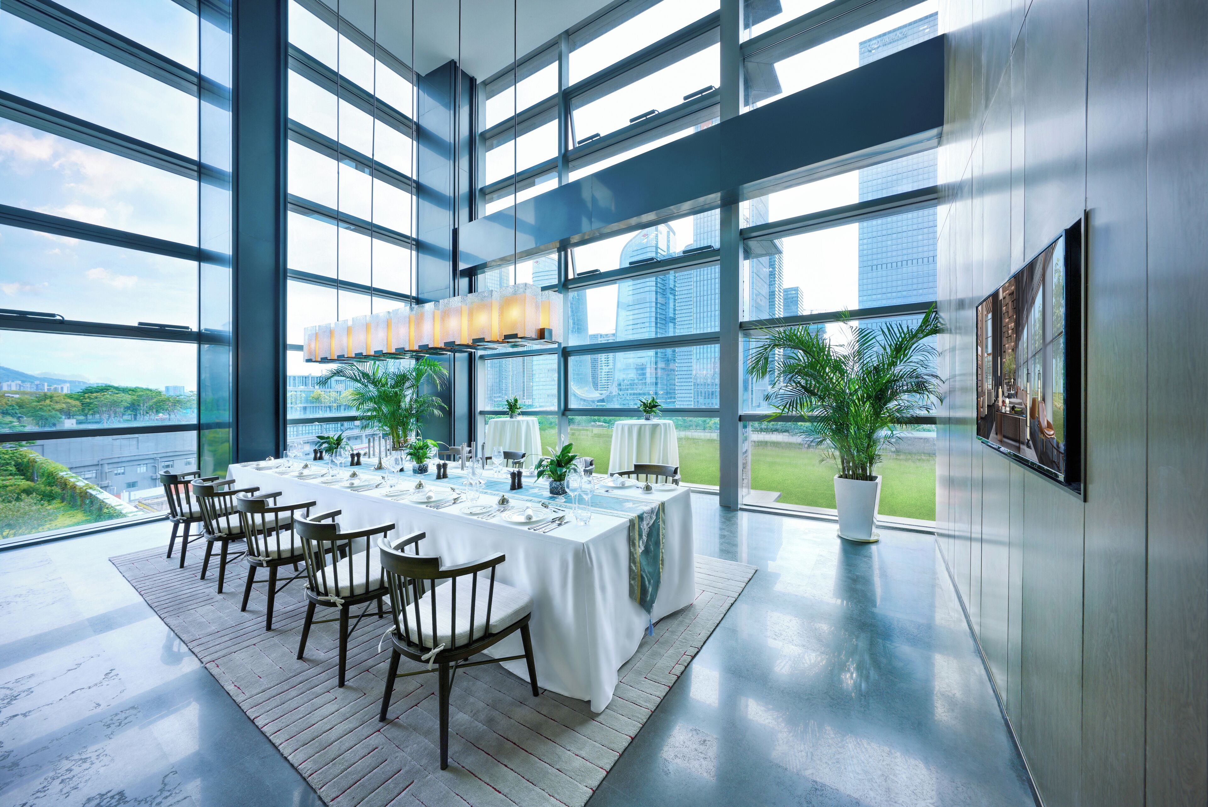 Uma sala de jantar moderna com uma mesa grande posta para refeição, janelas amplas com vista para a cidade e plantas internas.