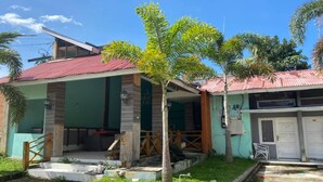 Um edifício de dois andares com telhado vermelho, varanda coberta e um pequeno jardim com uma palmeira.