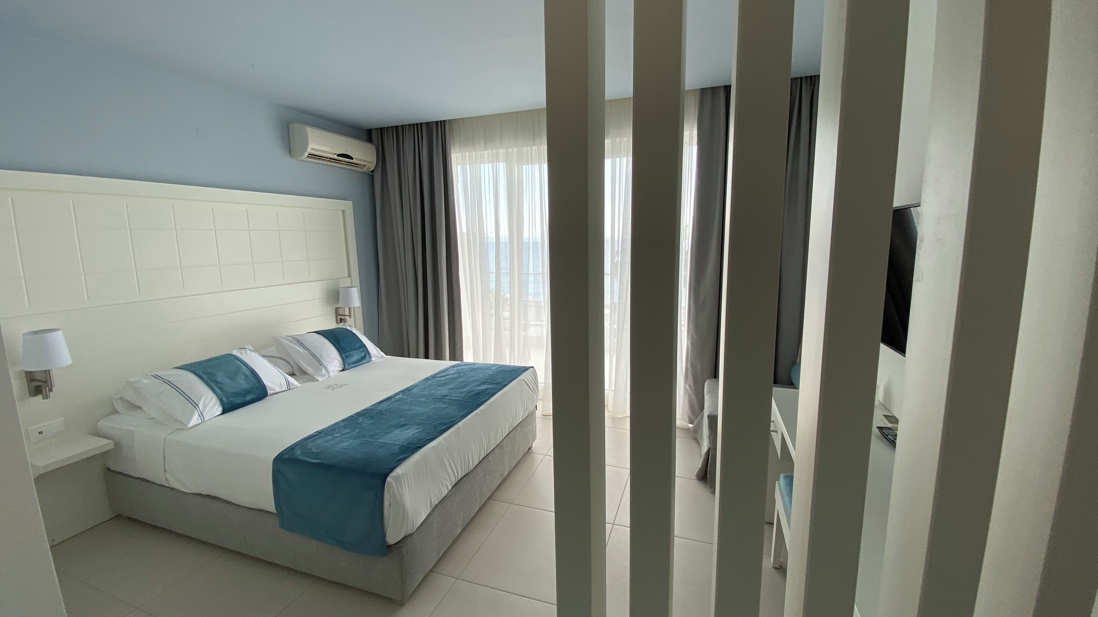 Quarto de hotel com uma cama grande, televisão de tela plana, janela com cortinas e um aparelho de ar condicionado na parede.