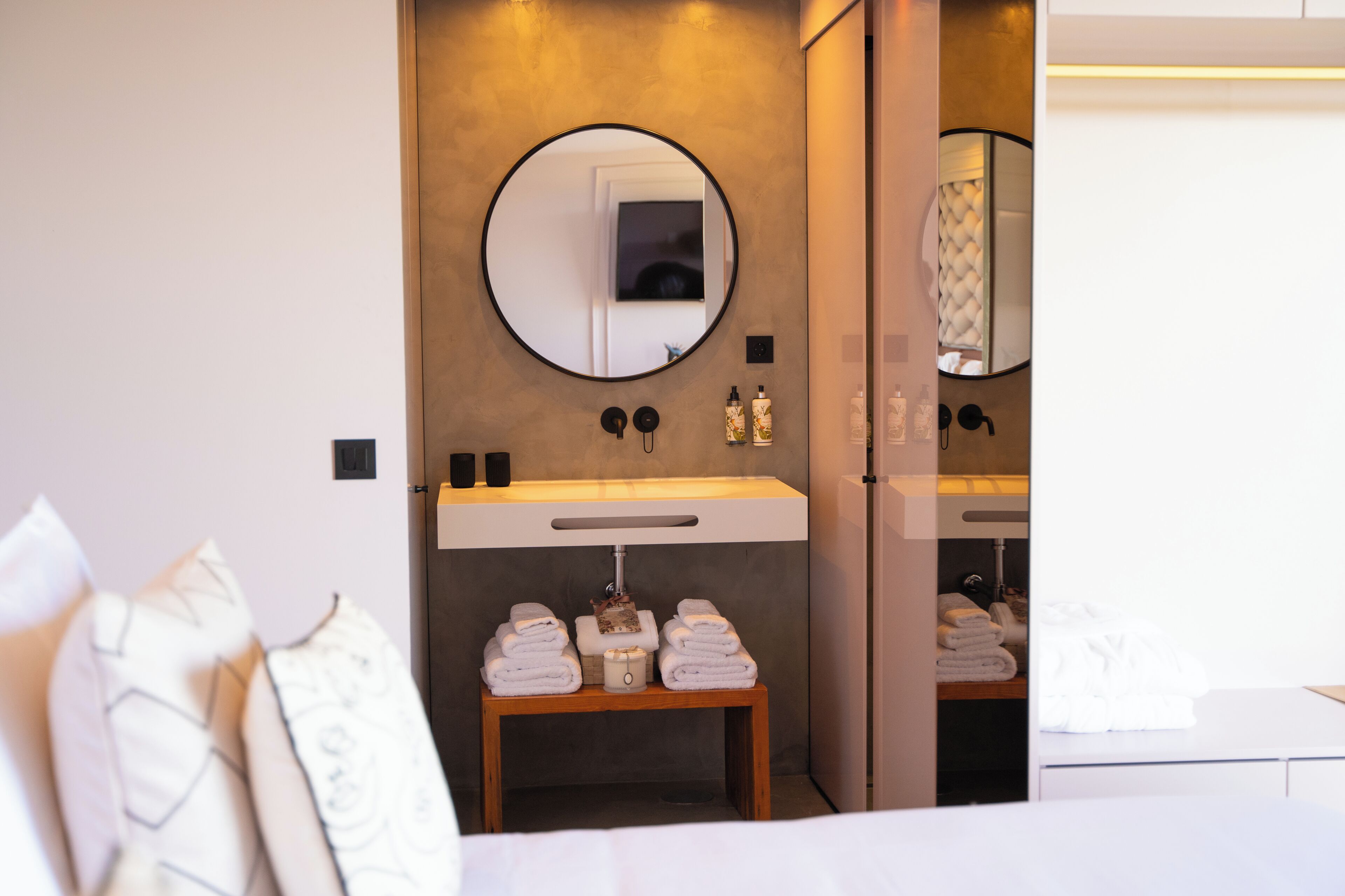 Quarto de hotel com um espelho redondo, um banco de madeira com toalhas e um banheiro moderno com um espelho grande.