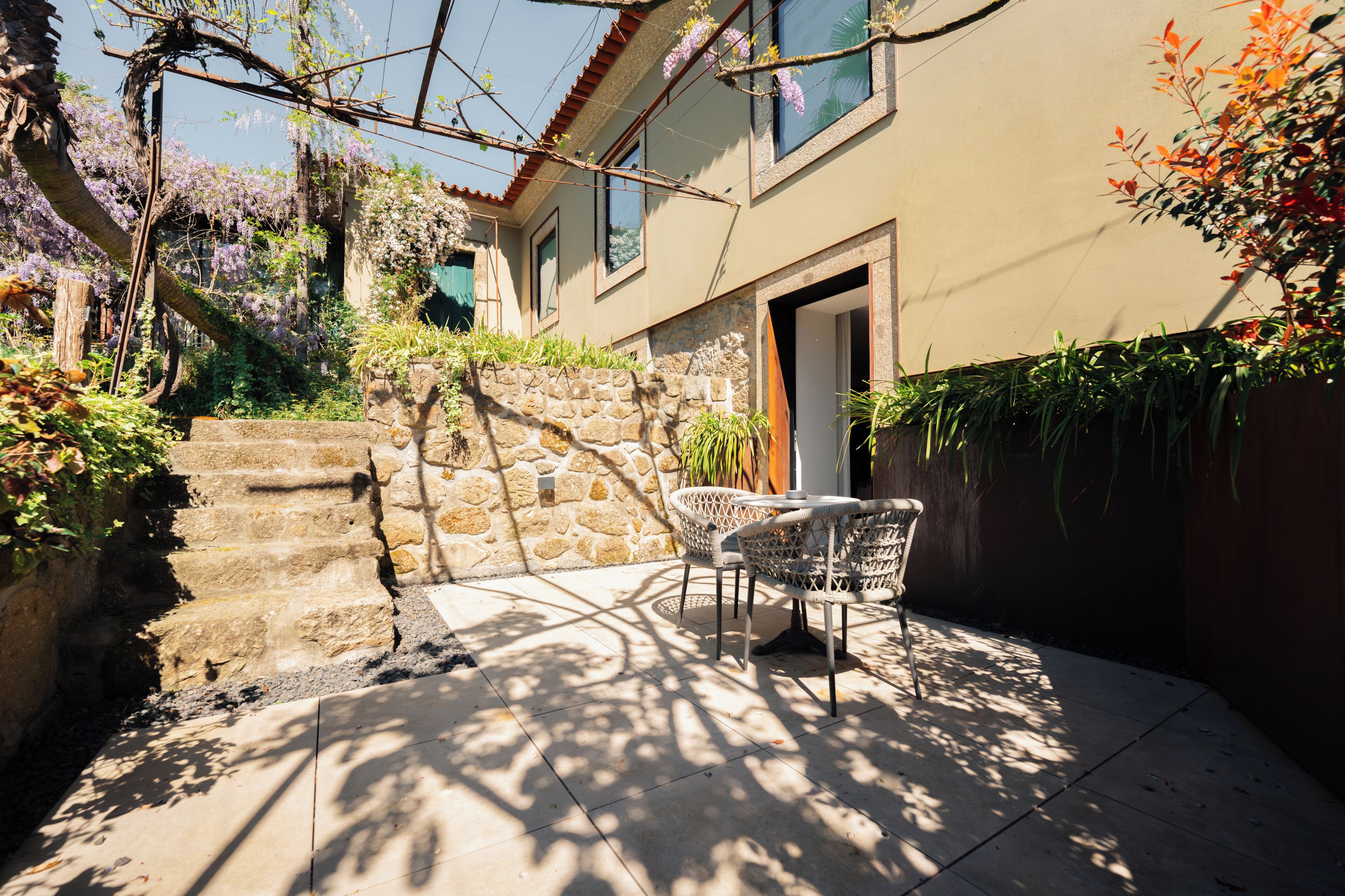 Escadaria de pedra que leva a uma casa com uma porta, uma pequena mesa ao ar livre com cadeiras e um jardim com diversas plantas.