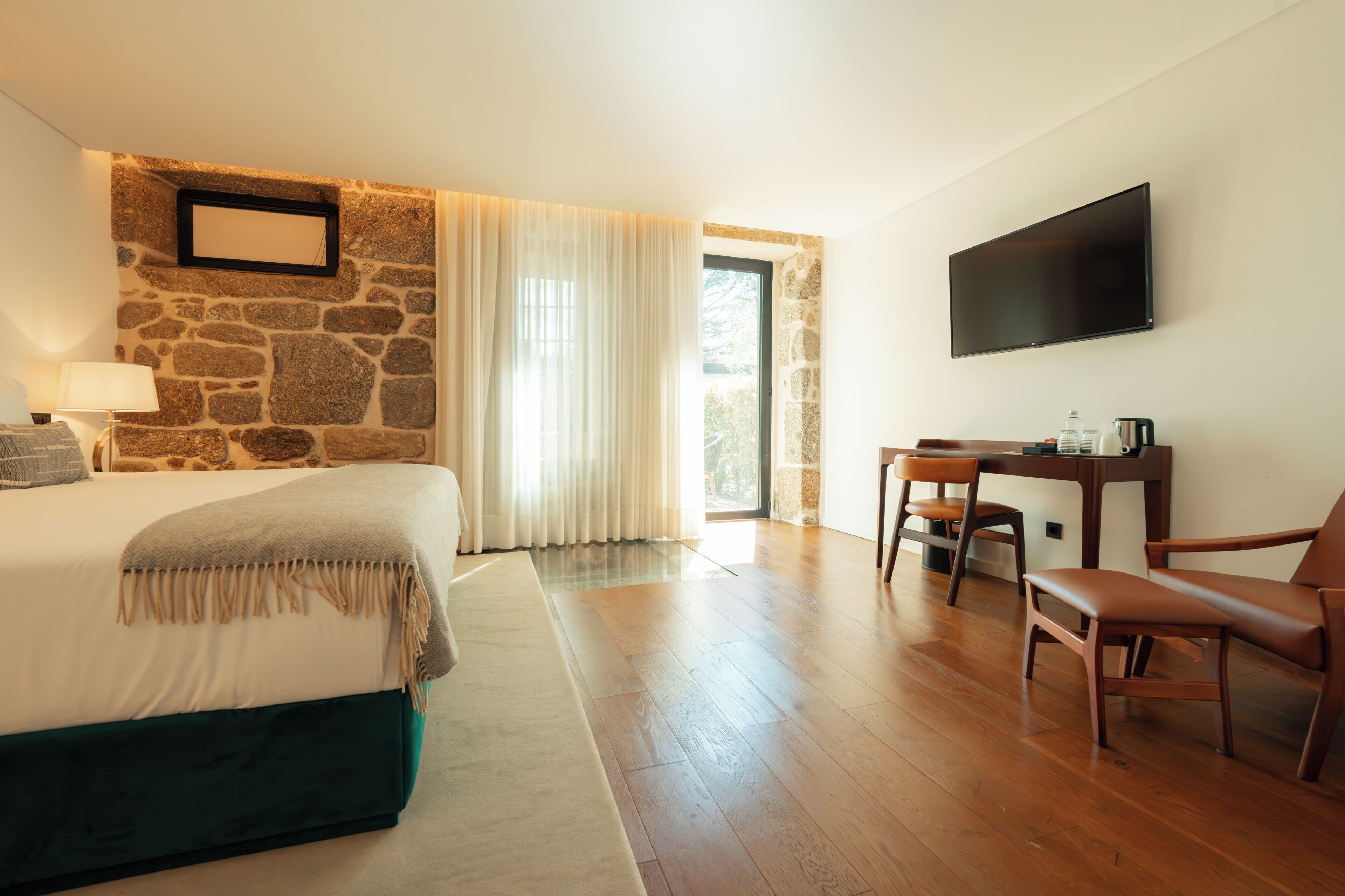 Quarto de hotel com uma cama, uma escrivaninha com cadeira, televisão e parede de pedra.