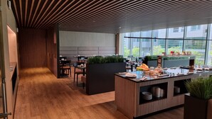 Um restaurante moderno com piso de madeira, uma ilha central com bancada preta e vista para o verde através de grandes janelas.