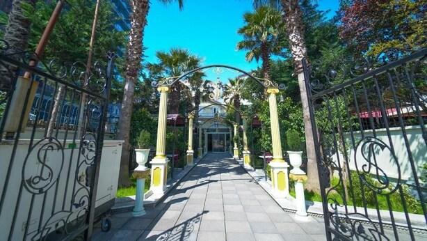 Uma passagem que leva à entrada de um edifício, com um arco decorativo, cercada por palmeiras e uma cerca de metal.