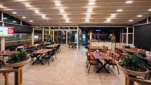 Uma ampla área de refeições interna com várias mesas e cadeiras, um bar e uma tela grande exibindo informações.