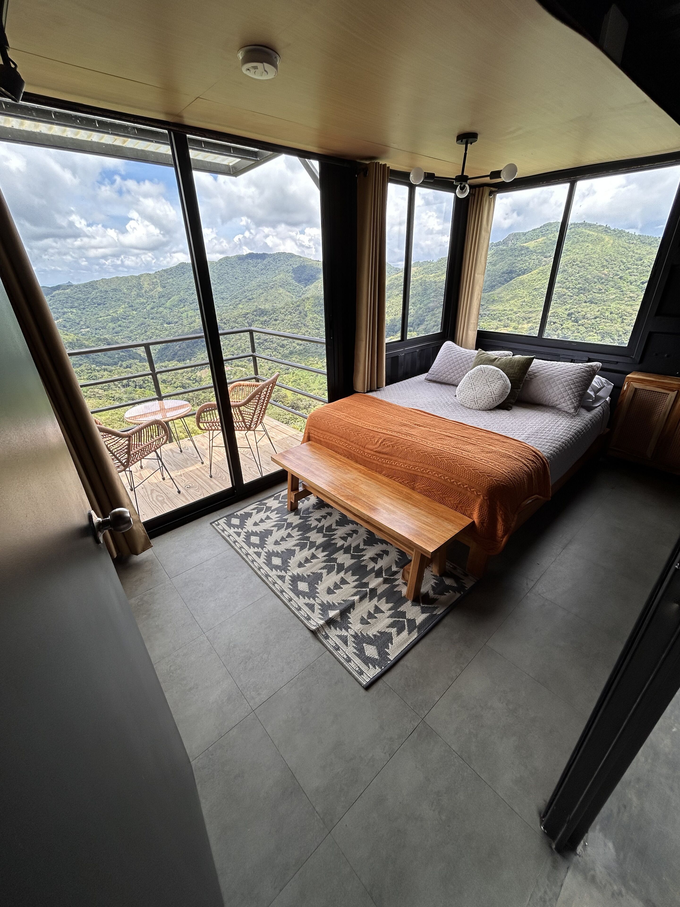 Quarto com uma cama grande, um banco de madeira, uma mesa pequena e vista para uma paisagem montanhosa através de grandes janelas.