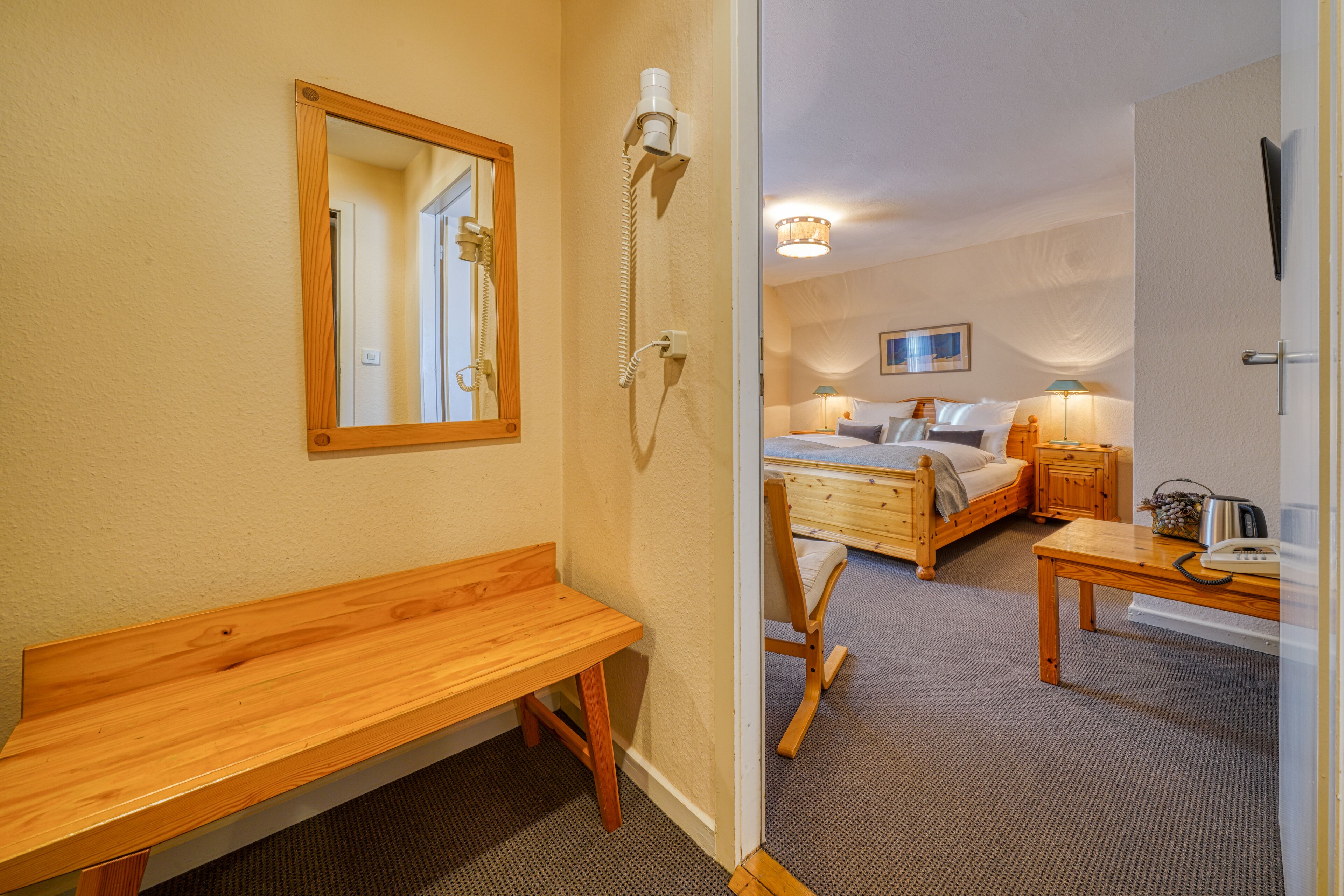 Quarto de hotel com um banco de madeira, uma cama com cabeceira, uma mesa de cabeceira com abajur e um espelho na parede.