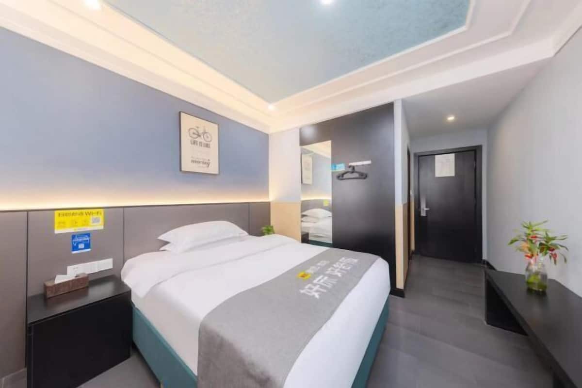 Quarto de hotel com cama, mesa de cabeceira, placa na parede, porta e uma pequena planta em cima de uma mesa.
