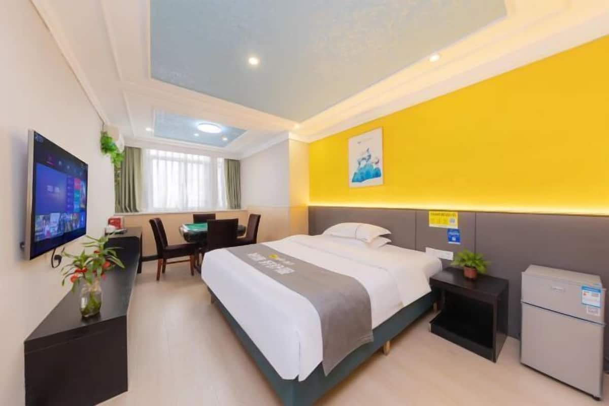 Quarto de hotel com cama, escrivaninha, cadeira, televisão e um pequeno frigorífico.