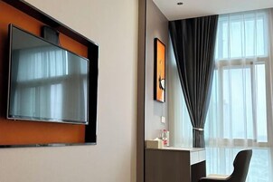Quarto de hotel moderno com uma TV de tela plana grande presa na parede, uma escrivaninha com cadeira e uma janela com cortinas translúcidas.