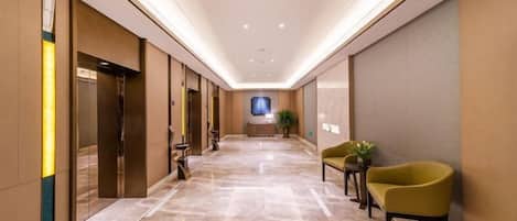 Corredor de hotel com piso de mármore, elevadores, balcão de recepção e uma cadeira verde.