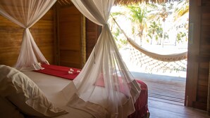 Quarto com cama coberta por um dossel branco, colcha vermelha e vista para palmeiras pela janela.