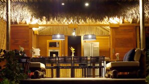 Um restaurante com telhado de palha, paredes de madeira e uma área de jantar central com cadeiras pretas e uma mesa.