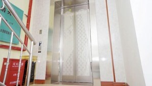 Um elevador com acabamento metálico e porta de vidro, situado num corredor com chão em mosaico e corrimão à esquerda.