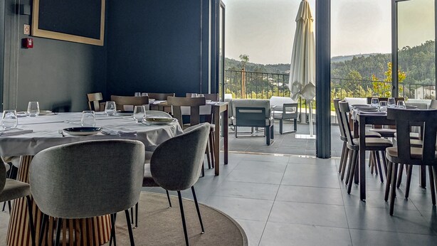 Restaurante com interior moderno, teto alto com vigas aparentes, grandes janelas com vista para o exterior e área de jantar com mesas e cadeiras.