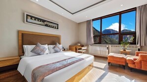 Quarto de hotel moderno com uma cama grande, área de estar com duas cadeiras, vista para montanhas através de amplas janelas e um painel decorativo na parede.