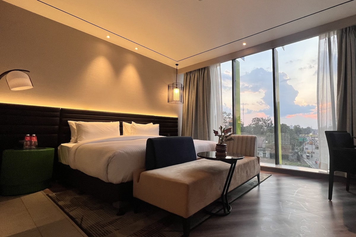 Quarto de hotel com uma cama grande, um sofá, uma escrivaninha e vista da cidade através de amplas janelas.