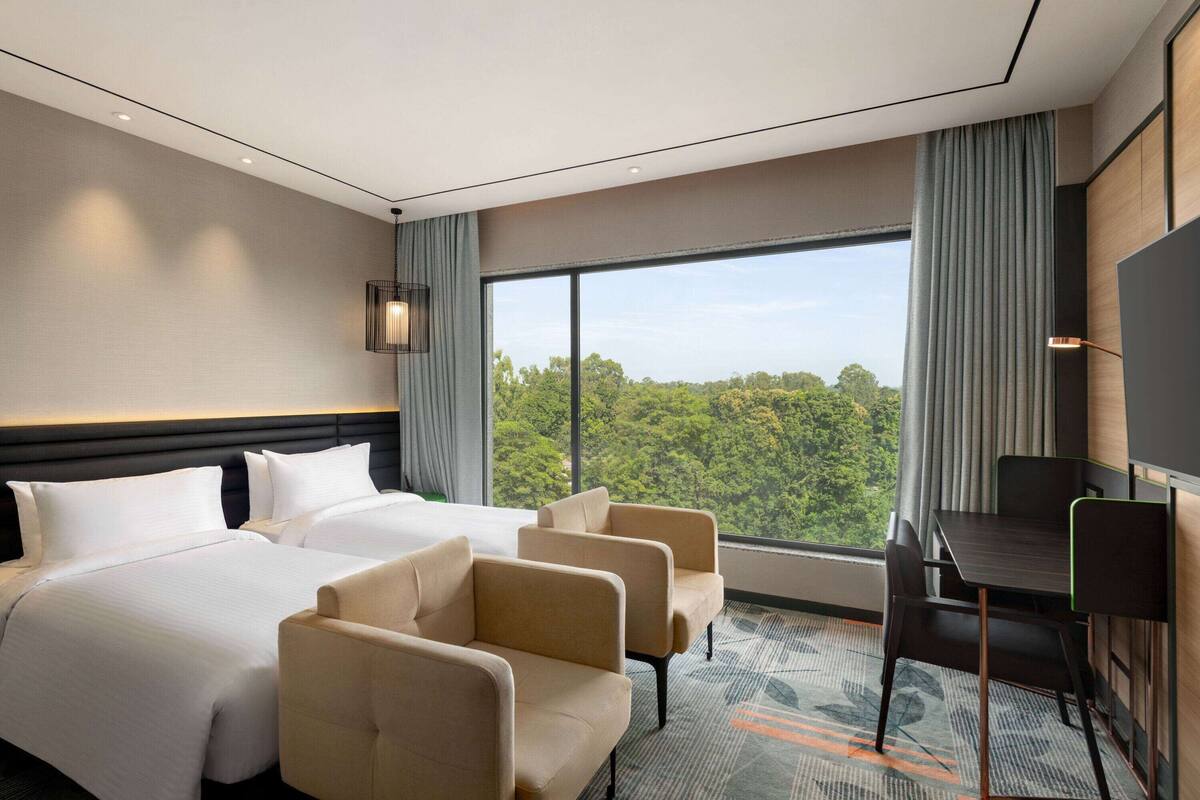 Quarto de hotel com uma cama grande, duas poltronas, uma mesa de escritório com cadeira, televisão e uma janela ampla com vista para árvores.