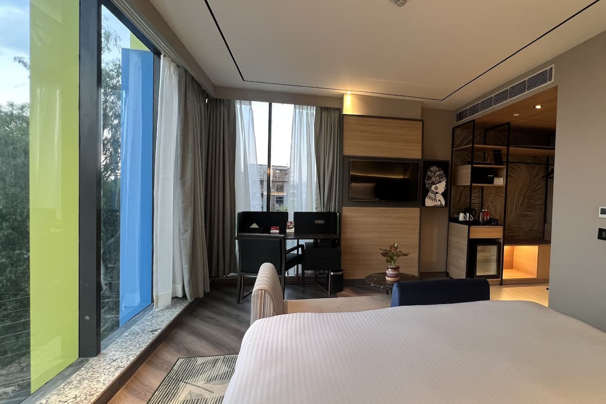 Quarto de hotel moderno com uma cama grande, uma mesa com cadeira, uma mesinha com planta e uma TV fixada na parede.