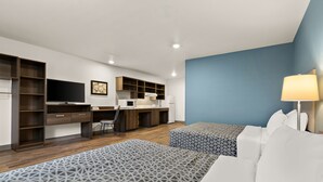 Quarto de hotel moderno com duas camas, uma escrivaninha, televisão e cozinha compacta.