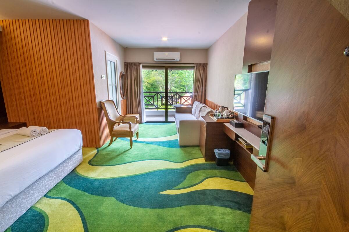 Quarto de hotel com cama, escrivaninha com cadeira, televisão e varanda com vista para o verde.