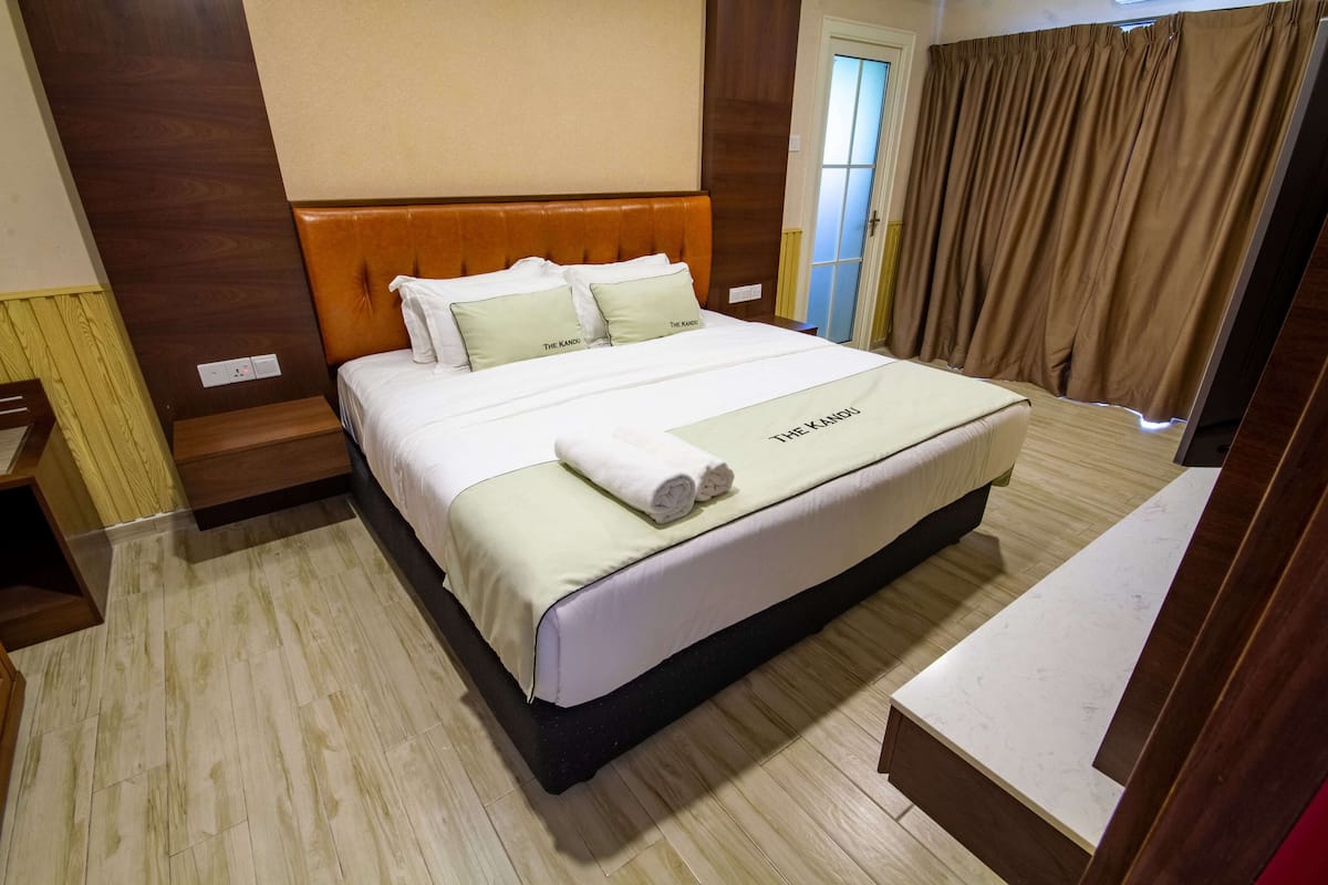 Quarto de hotel com uma cama, mesa de cabeceira, cabeceira de madeira e vista para o exterior através de uma janela.