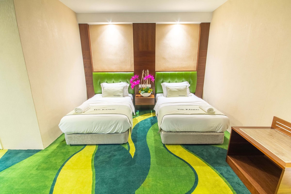 Quarto de hotel com duas camas, cabeceira verde, uma mesa pequena e um tapete colorido.