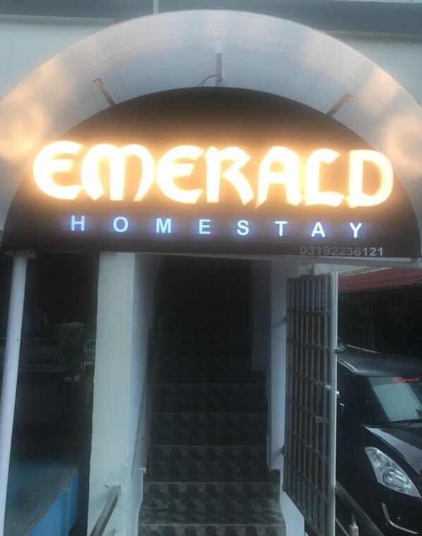 Entrada de hotel com um letreiro de neon que diz 'Emerald Homestay' e um número de contato.