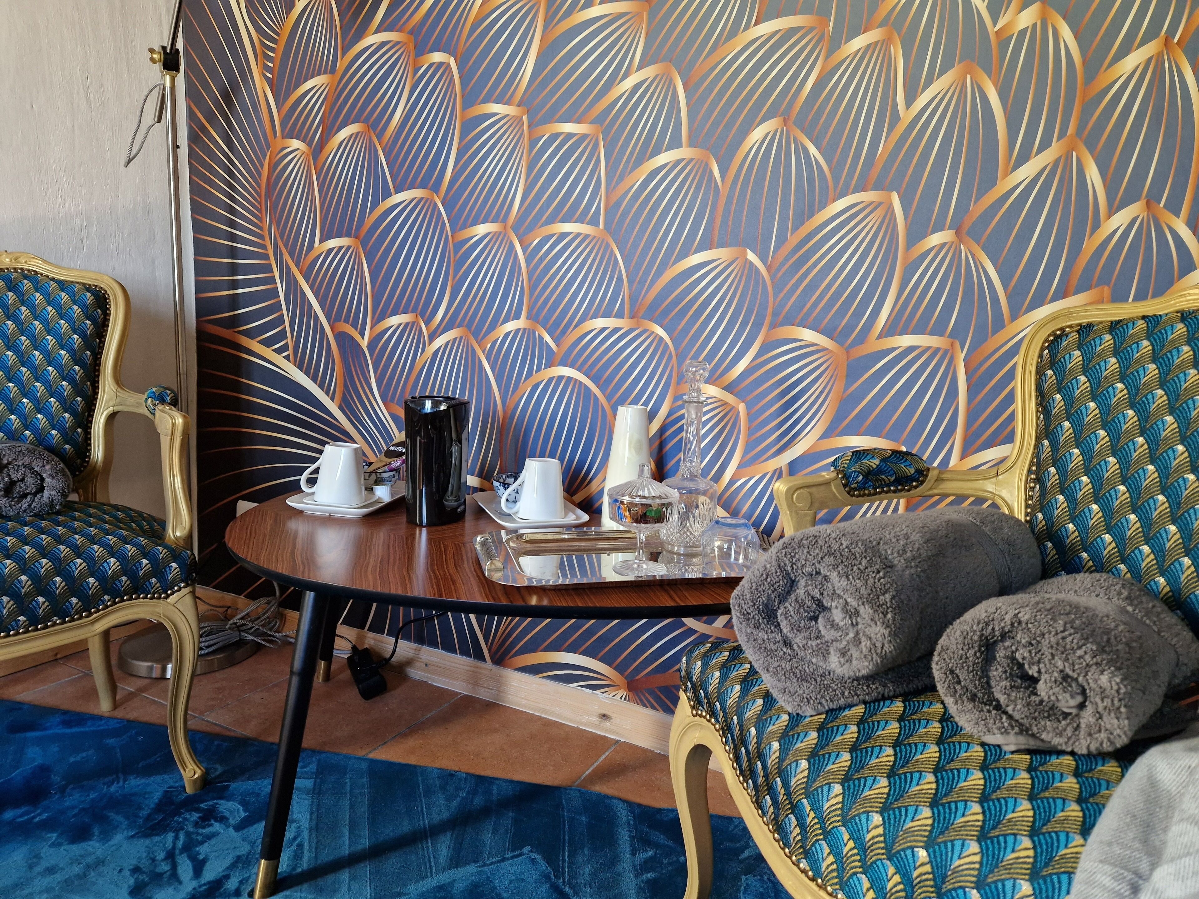 Um quarto com papel de parede estampado, duas poltronas estofadas, uma mesa de madeira redonda com um tabuleiro e um tapete azul.
