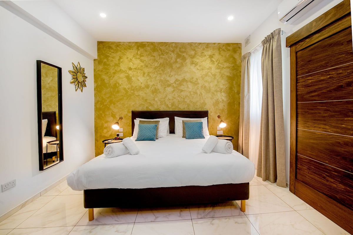Uma cama grande com roupa de cama branca, duas mesas de cabeceira com abajures, um espelho e um guarda-roupa de madeira.