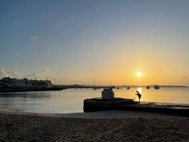 Uma praia com alguém pescando perto de um quebra-mar, barcos na água e edifícios ao fundo.