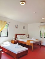 Quarto de hotel com duas camas, uma cabeceira de madeira, mesa de cabeceira com abajur, janela com cortinas e um aparelho de ar condicionado.